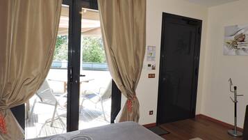 Quarto de hotel com cama, porta, janela com cortinas, uma cadeira e uma mesa na varanda.