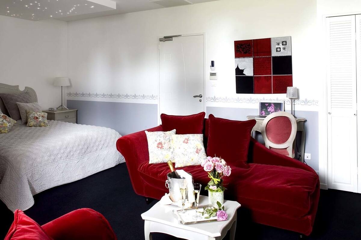Um quarto com uma cama, um sofá vermelho, uma mesinha com um vaso de flores e um espelho.