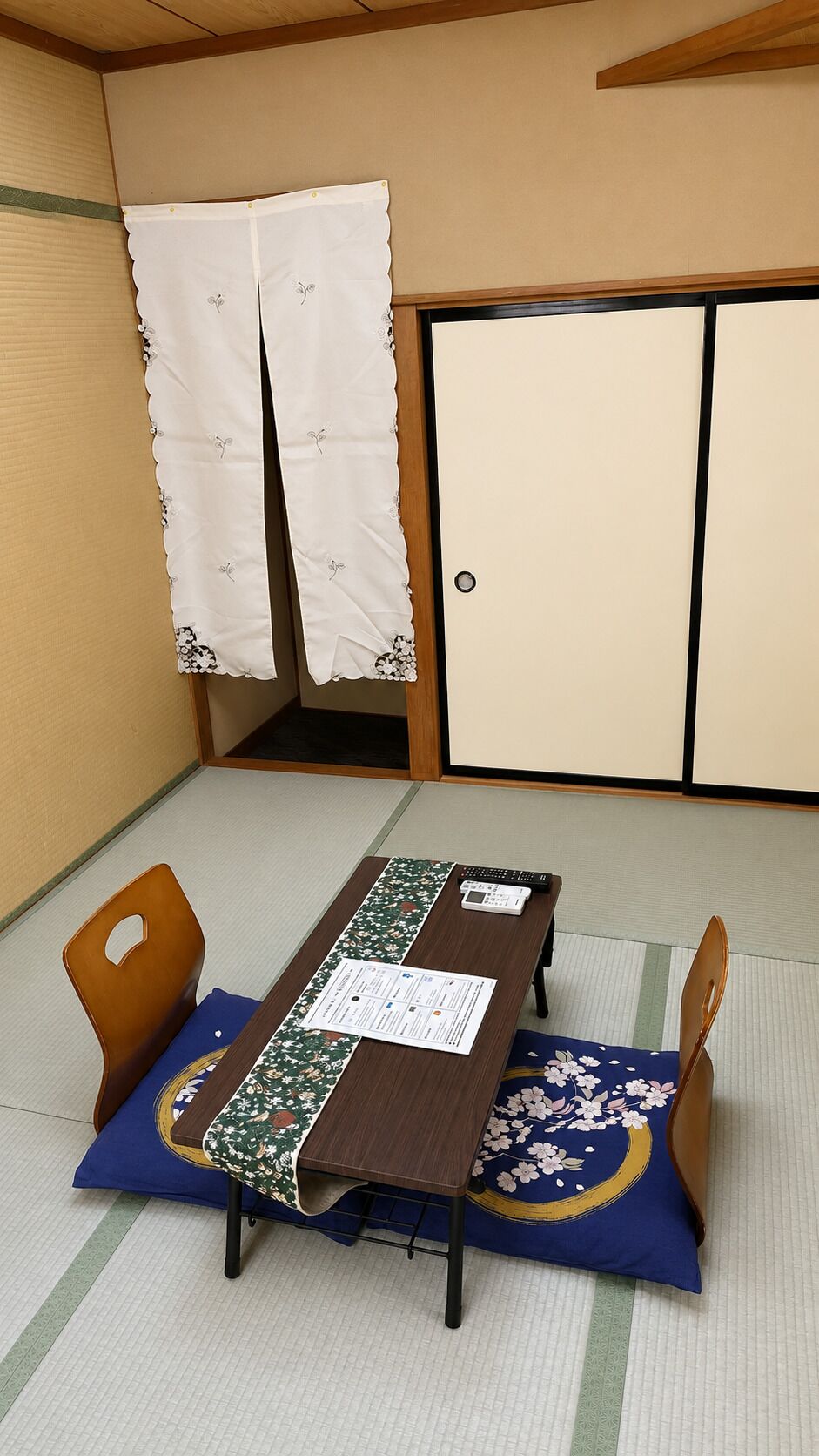 Um quarto japonês tradicional com tatames, uma mesa baixa com um jogo de mesa floral, duas cadeiras de madeira e uma porta deslizante com cortina branca.