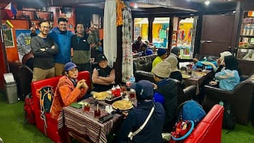 Uma área de refeições com mesas e cadeiras, pessoas sentadas a comer e um balcão com produtos à venda.