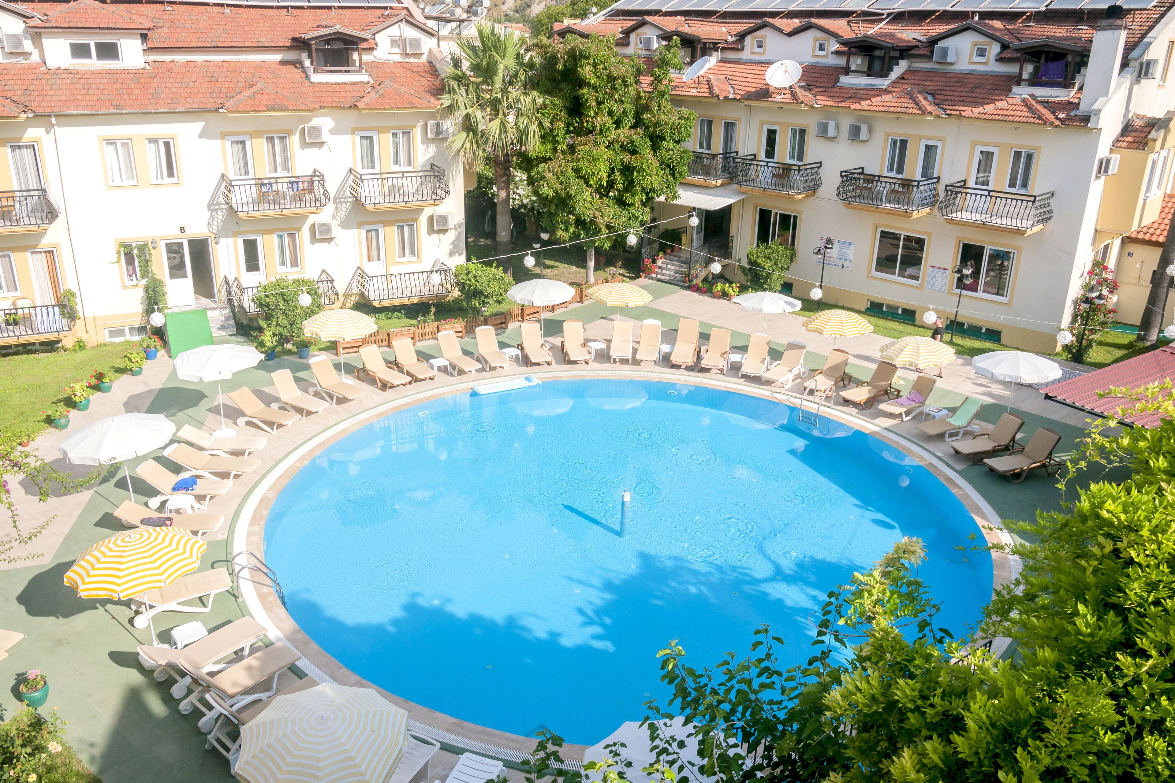 Uma piscina circular com espreguiçadeiras e guarda-sóis, rodeada por um edifício com varandas e um jardim.