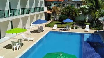 Piscina com espreguiçadeiras e guarda-sóis, cercada por um edifício com amplas janelas e vista para palmeiras.
