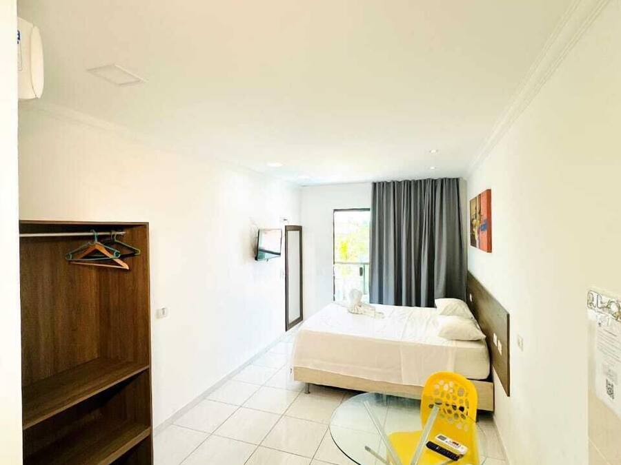 Quarto de hotel com cama, guarda-roupa de madeira, mesa de vidro com cadeira e uma pequena mesa com cadeira.