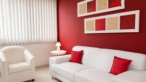 Uma sala de estar com um sofá branco, duas almofadas vermelhas, uma poltrona branca e uma pequena mesa branca com um abajur.
