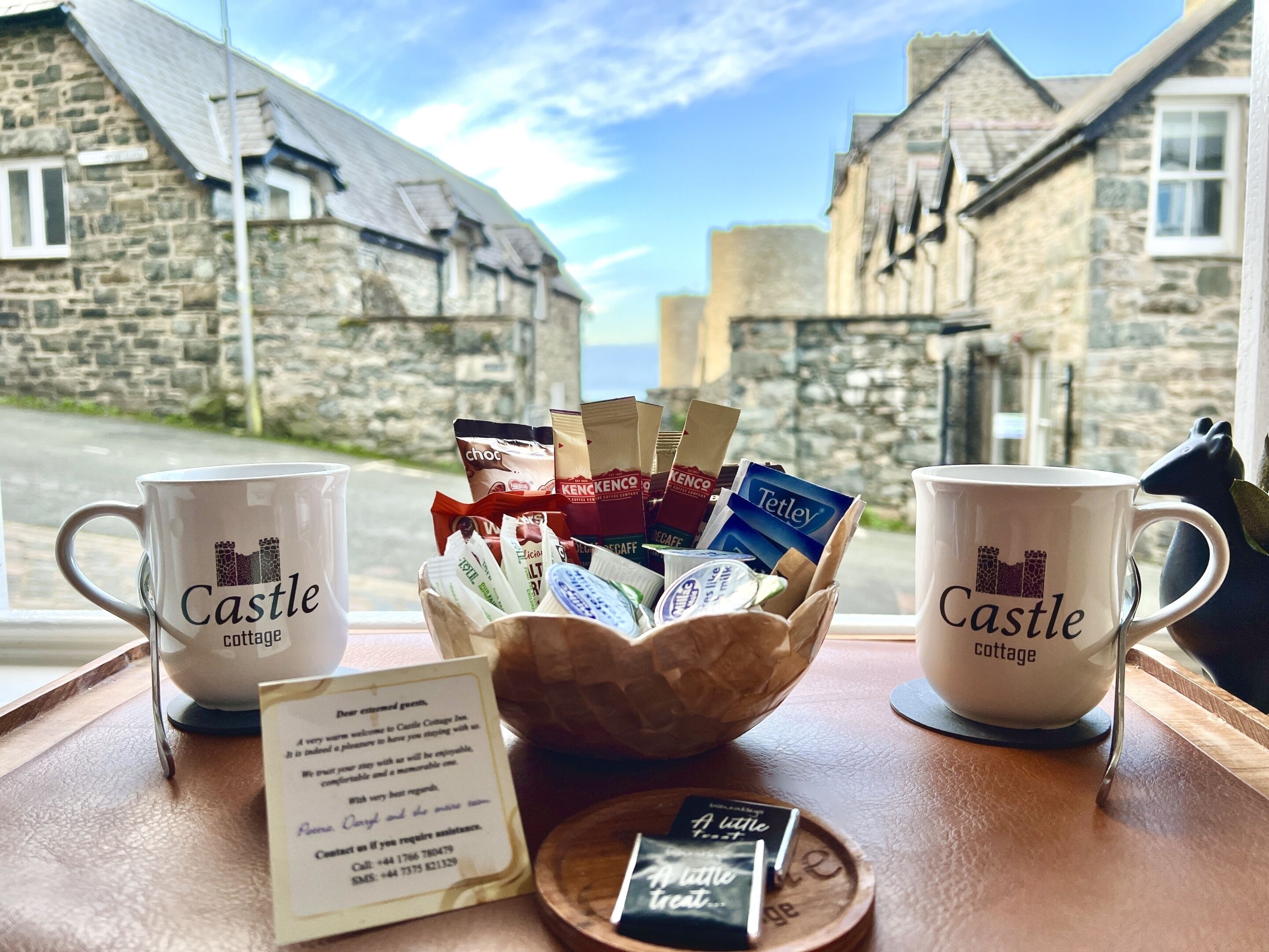 Duas canecas com a marcação "Castle Cottage", uma tigela de madeira cheia de várias barras de chocolate e um postal de boas-vindas sobre um apoio de madeira.