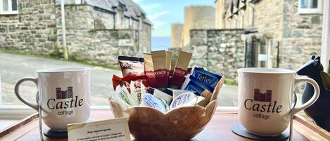 Duas canecas com a marcação "Castle Cottage", uma tigela de madeira cheia de várias barras de chocolate e um postal de boas-vindas sobre um apoio de madeira.