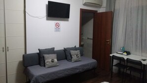 Um quarto pequeno com um sofá, uma televisão na parede, uma mesa com cadeiras e uma porta.