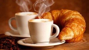 Duas chávenas de café fumegante, um croissant e grãos de café sobre uma superfície de madeira.