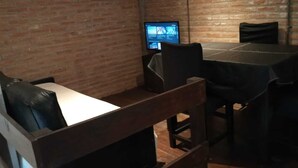 Um quarto com paredes de tijolo, uma mesa, cadeiras, uma televisão na parede e um banco.