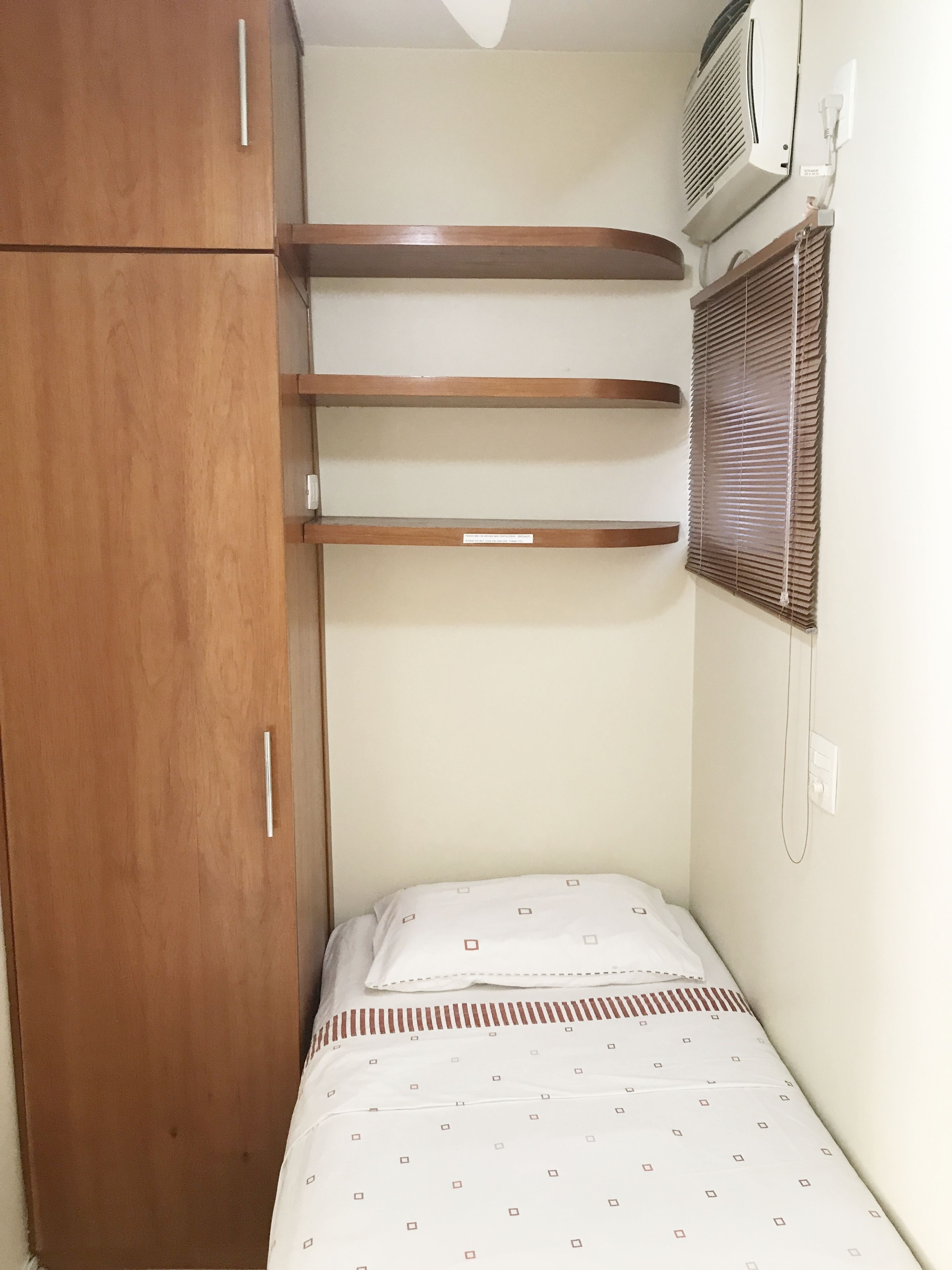 Quarto pequeno com uma cama de solteiro, guarda-roupa de madeira, ventoinha no teto e janela com persianas.