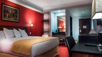 Quarto de hotel com uma cama grande, uma escrivaninha com cadeira, uma mesinha de cabeceira com abajur e um quadro na parede.