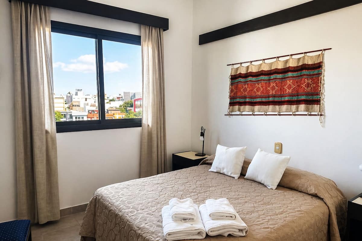 Quarto de hotel com cama, duas almofadas, janela com cortinas, tapeçaria na parede e mesa de cabeceira com abajur.