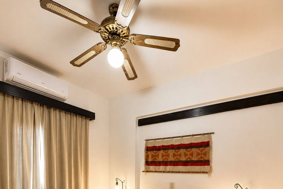 Quarto de hotel com cama, ventoinha no teto, duas mesas de cabeceira com luminárias, ar-condicionado fixado na parede e janela com cortinas.