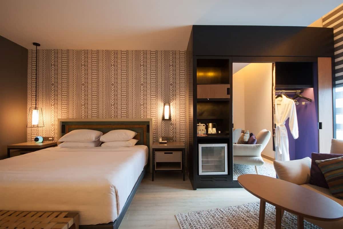 Quarto de hotel moderno com cama, mesas de cabeceira, área de estar com uma poltrona, pequena cozinha e closet espaçoso.