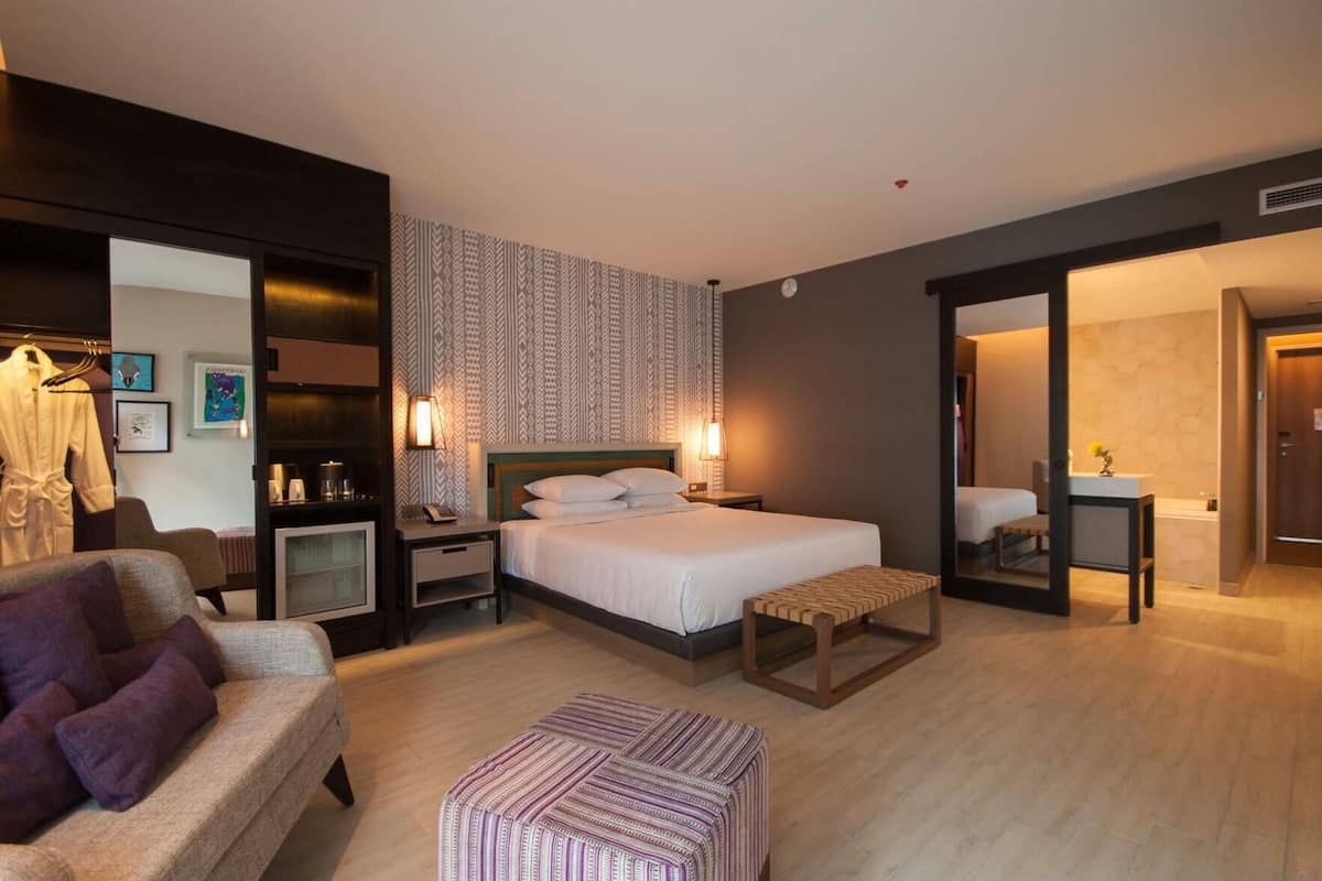 Quarto de hotel com cama, sofá, mesinha, espelho, banheiro e televisão.