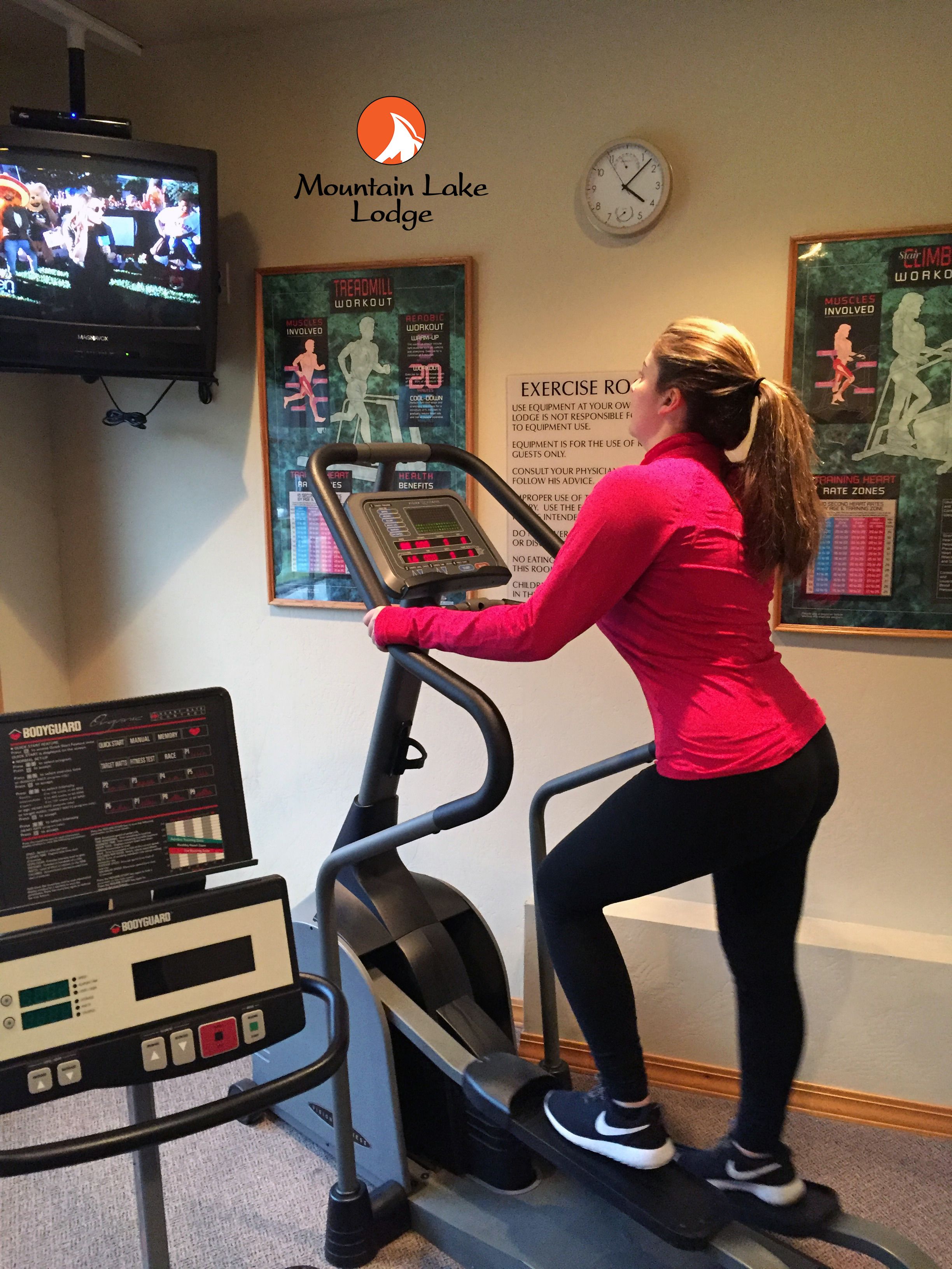 Mulher usando uma esteira numa academia, com uma televisão exibindo um evento esportivo, um relógio e cartazes de exercícios na parede.