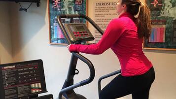 Mulher usando uma esteira numa academia, com uma televisão exibindo um evento esportivo, um relógio e cartazes de exercícios na parede.