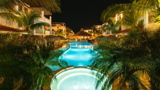 Vista noturna de uma piscina infinita em um resort, com palmeiras e estruturas de telhado de palha.