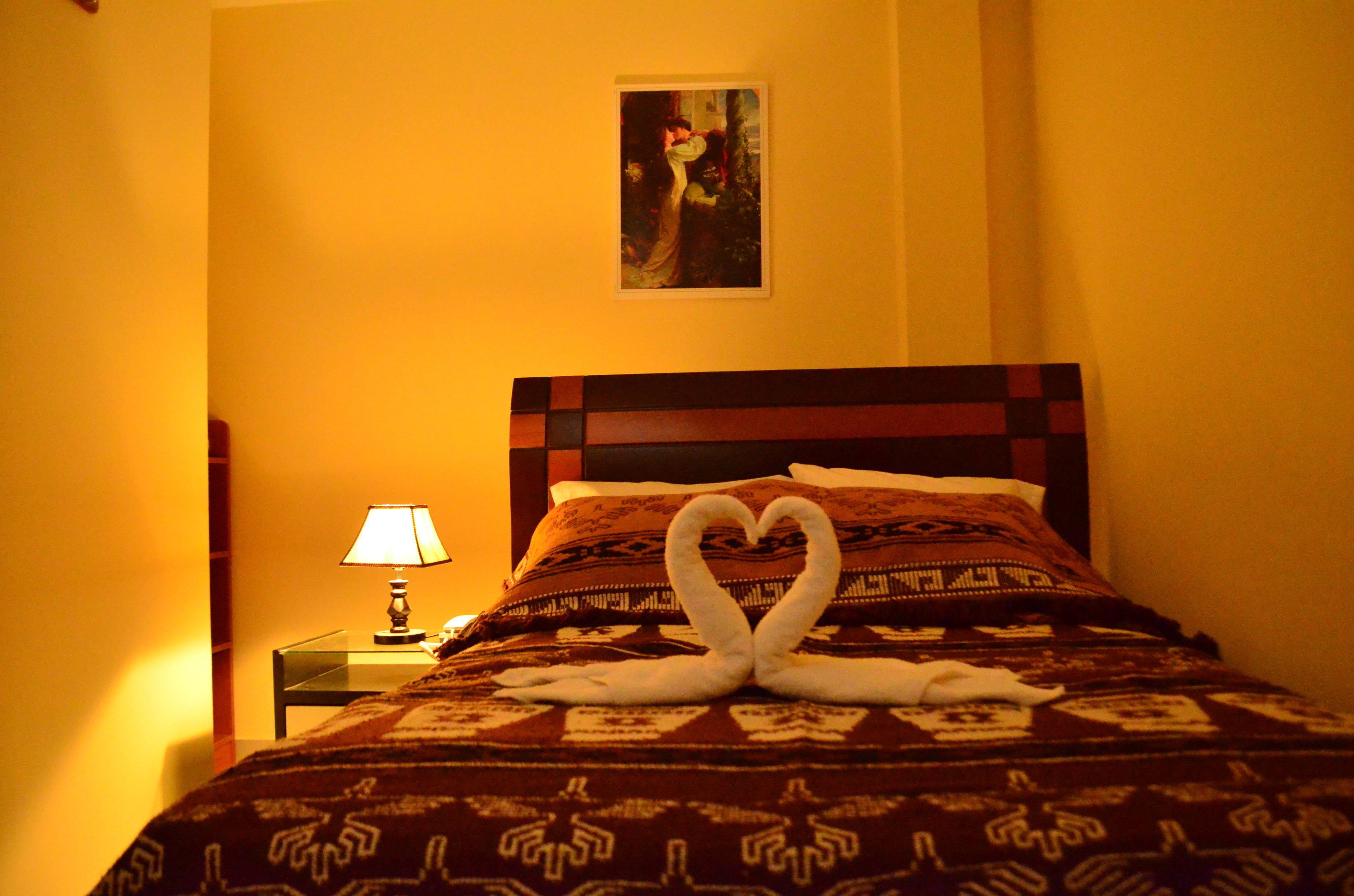 Quarto de hotel com uma cama, apresentação de toalha em formato de coração, mesa de cabeceira com abajur e um quadro na parede.