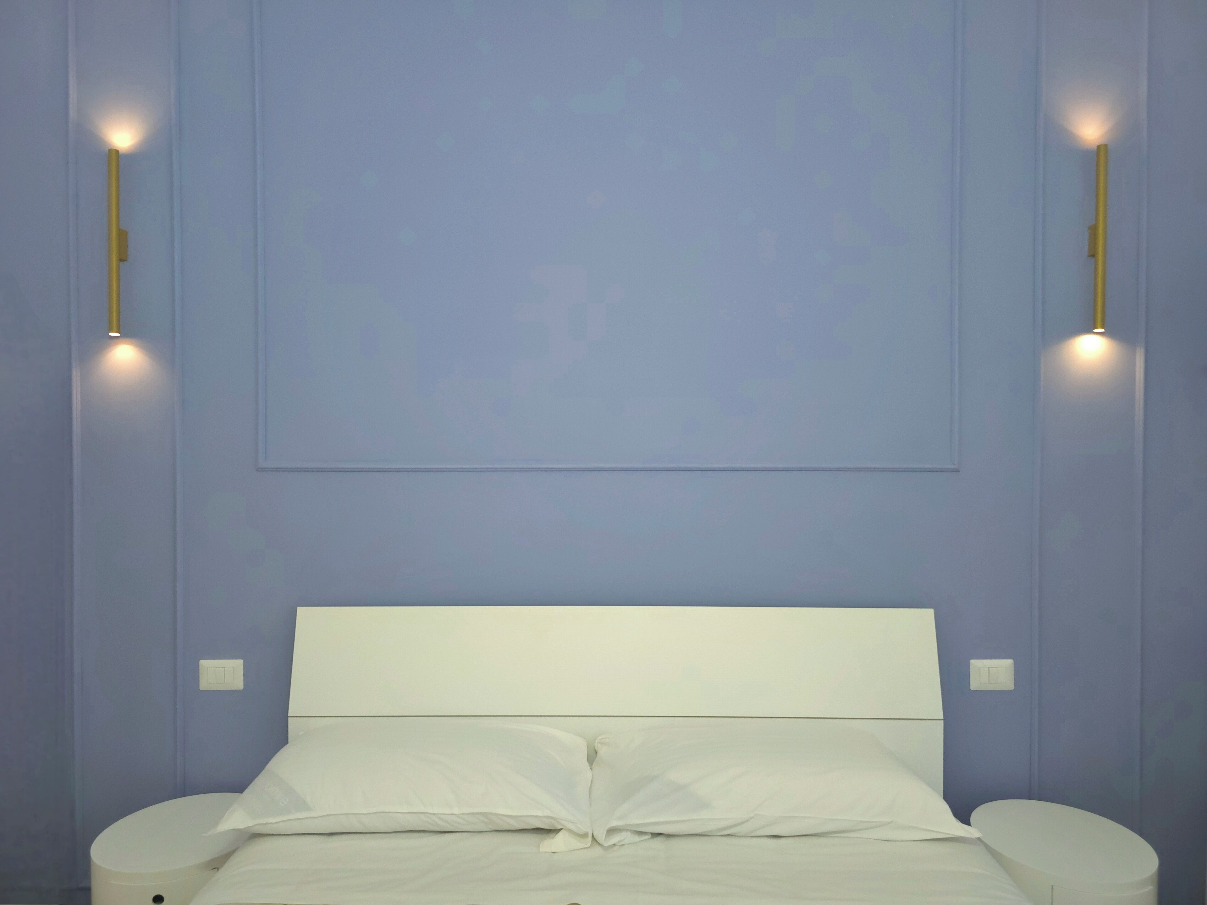 Cama com roupa de cama branca, duas mesas de cabeceira e luminárias fixadas na parede.