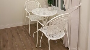 Um quarto pequeno com duas cadeiras de metal branco e uma mesa, uma planta em vaso e uma janela com cortinas brancas.