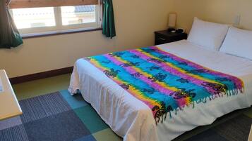 Quarto de hotel com uma cama grande, uma mesa de cabeceira com abajur, uma cadeira e uma janela com cortinas.