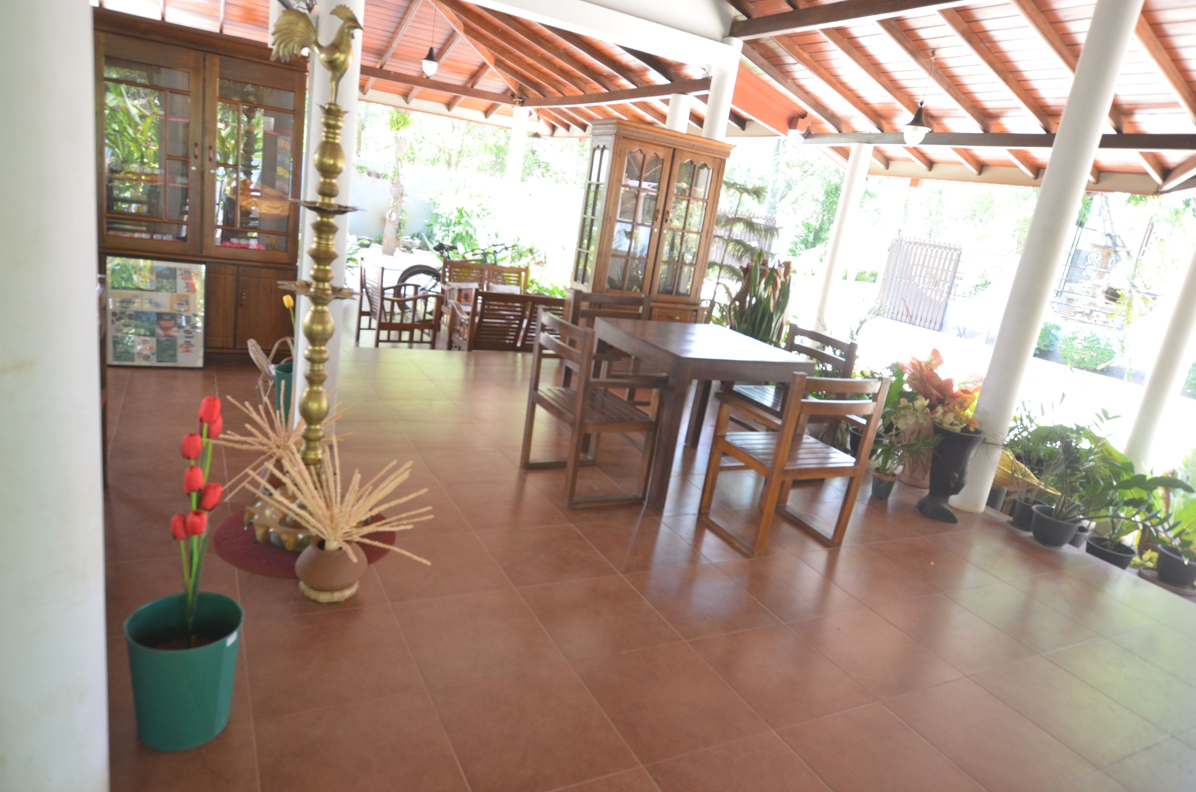 Uma área de varanda coberta com móveis de madeira, plantas em vasos e piso de azulejos.