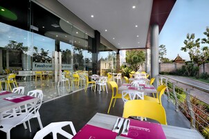 Uma área de refeições ao ar livre com cadeiras amarelas e brancas, mesas com cardápios rosa e um muro de vidro com a sinalização 'favehotel'.