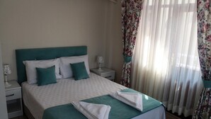 Uma cama bem arrumada com cabeceira azul-turquesa e almofadas, uma mesa de cabeceira branca com abajur, e cortinas florais em uma janela.
