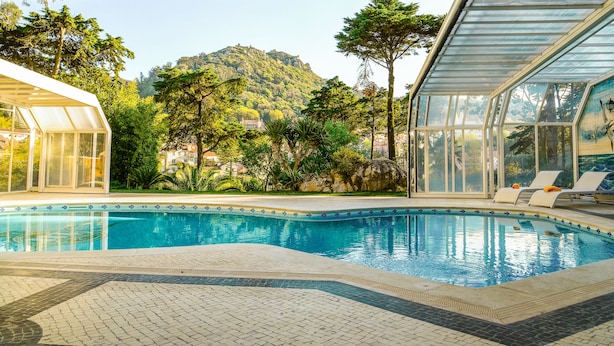 Piscina com água azul cristalina, rodeada por um deck ladrilhado e uma área de lounge envidraçada com cadeiras.