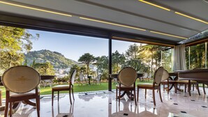 Um quarto espaçoso com uma grande janela que oferece vista para um jardim exuberante e uma montanha. Há várias cadeiras de madeira e um piano.