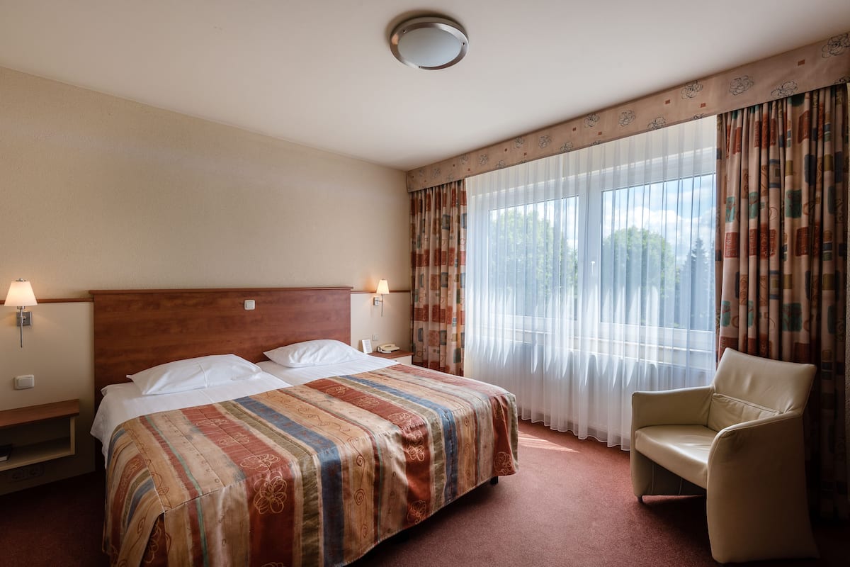 Quarto de hotel com uma cama grande, uma cadeira, um criado-mudo e uma janela com cortinas.
