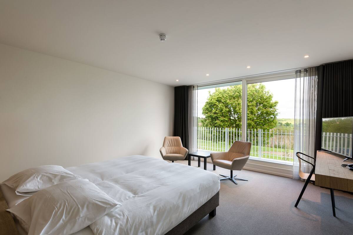 Quarto de hotel moderno com uma cama grande, duas poltronas, uma mesinha e uma televisão.