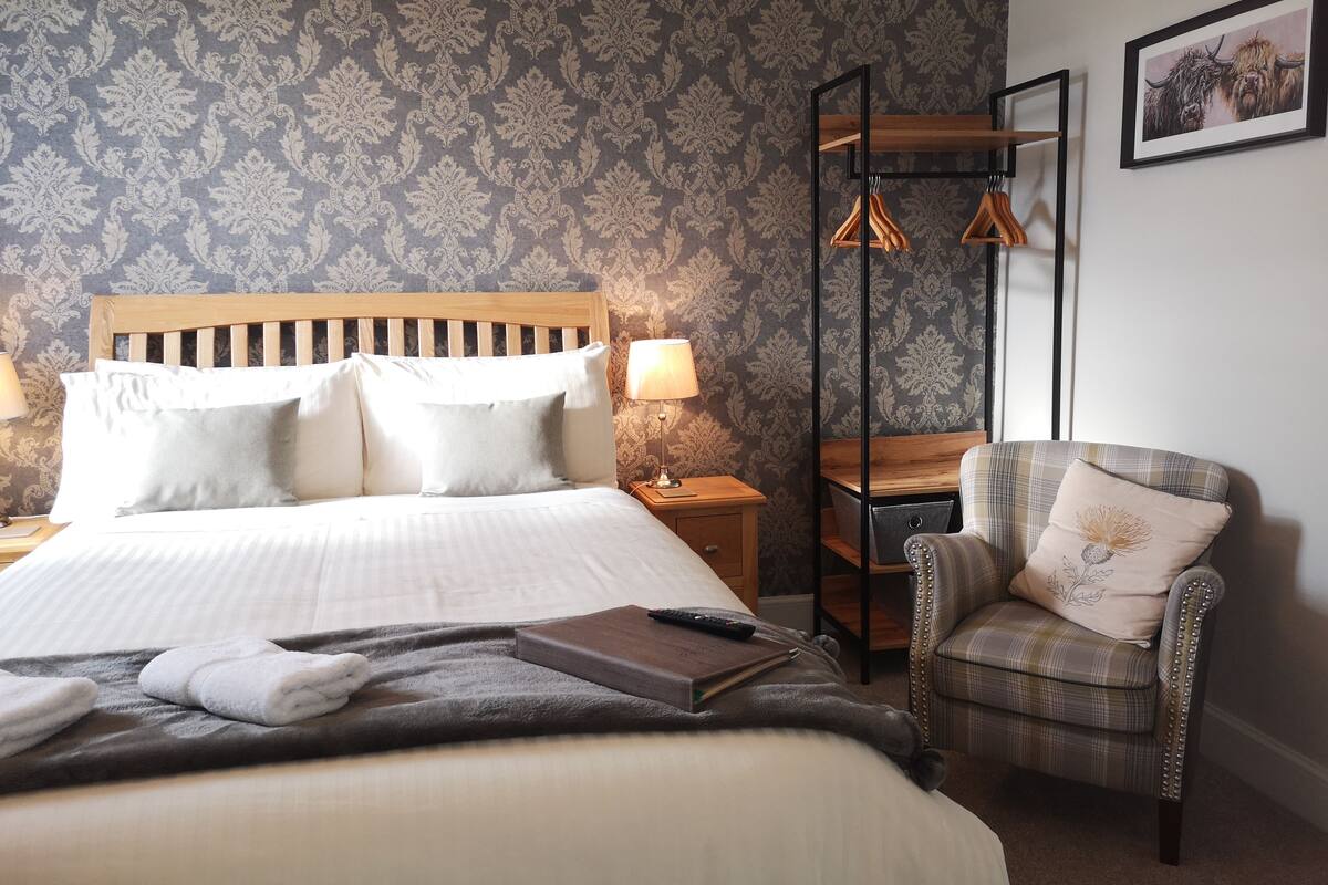 Cama bem arrumada com almofadas, mesa de cabeceira com abajur, poltrona xadrez e quadro na parede.
