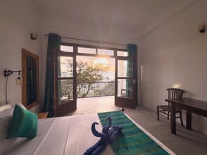 Quarto de hotel com cama, escrivaninha e varanda com vista para árvores e céu.