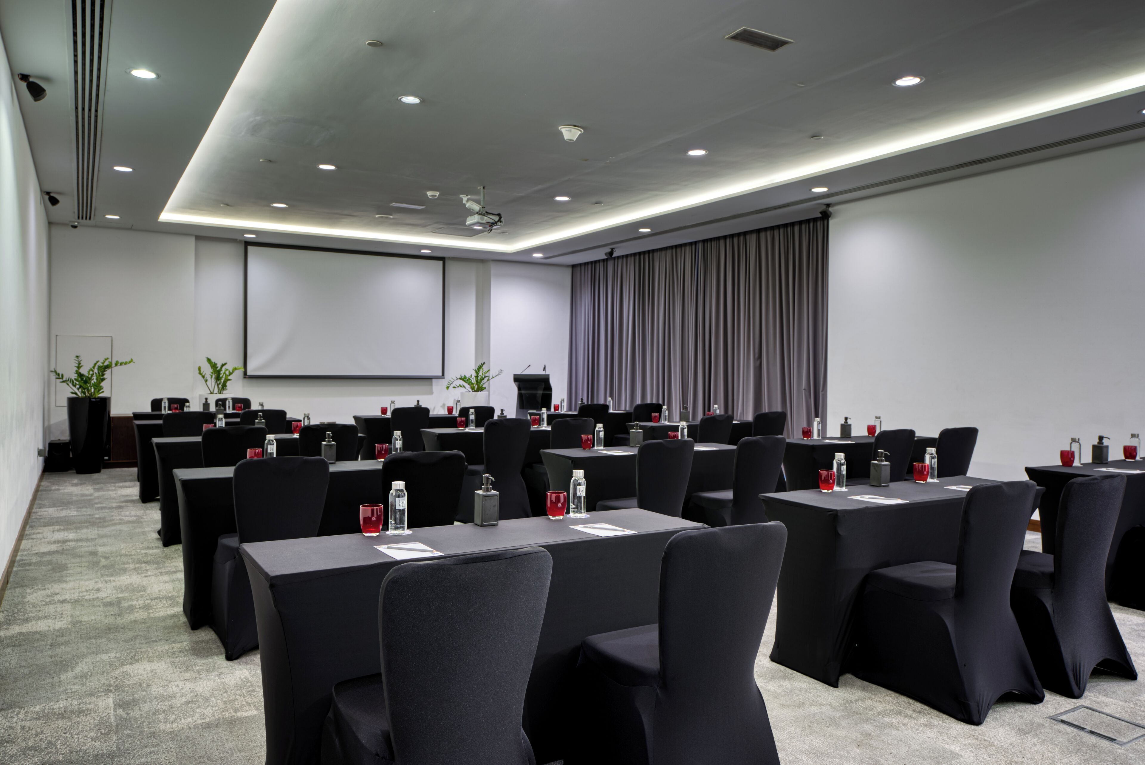 Uma sala de conferências com várias mesas e cadeiras, equipada com porta-nomes, garrafas de água e uma tela de projeção.