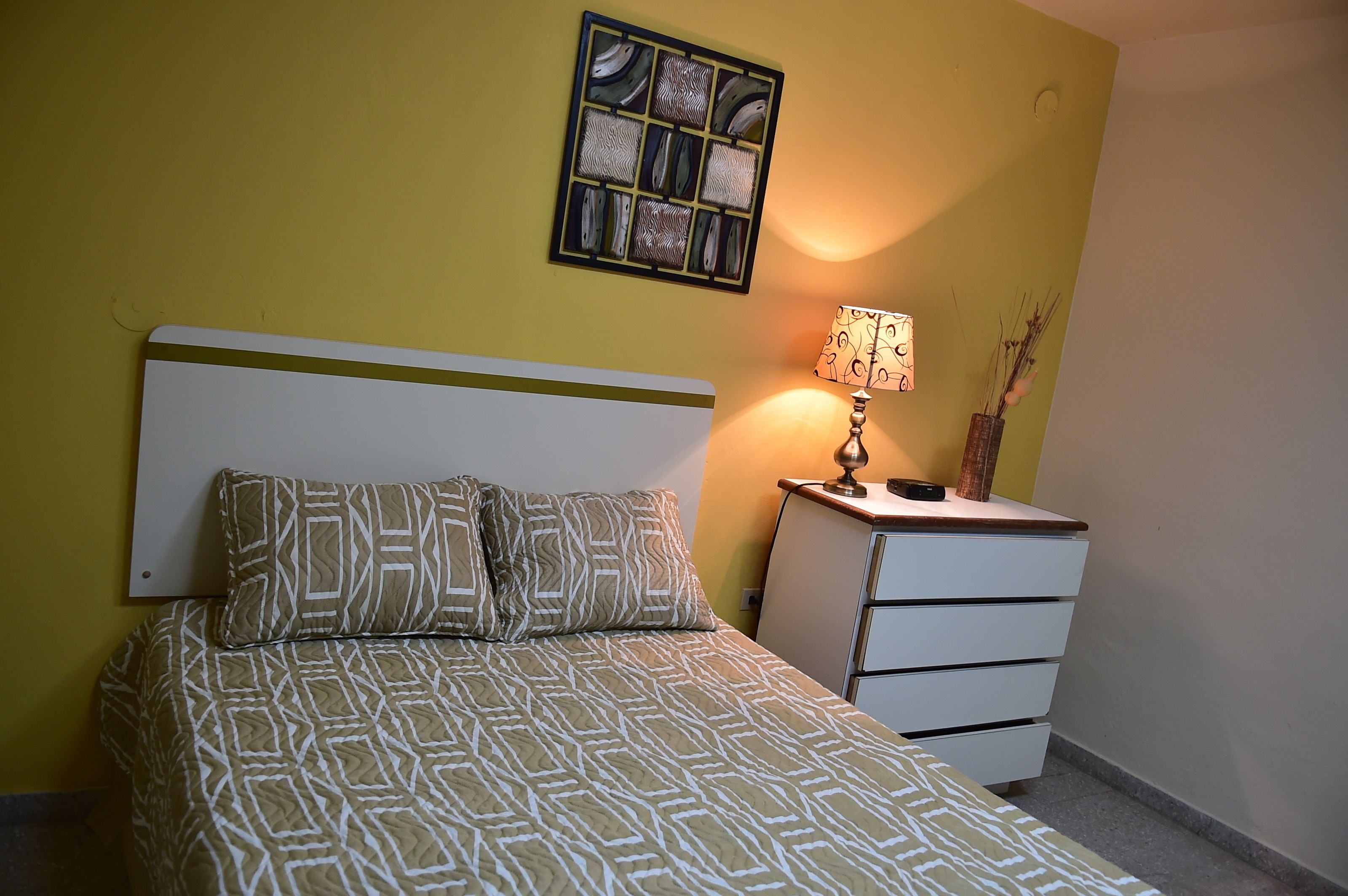 Uma cama com roupa de cama estampada, uma luminária de cabeceira, uma pequena mesa de cabeceira de madeira e um objeto decorativo na parede.