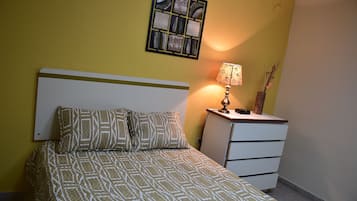 Uma cama com roupa de cama estampada, uma luminária de cabeceira, uma pequena mesa de cabeceira de madeira e um objeto decorativo na parede.