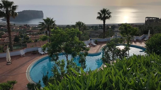 Uma área de piscina com espreguiçadeiras, palmeiras e vista para o mar e um penhasco.