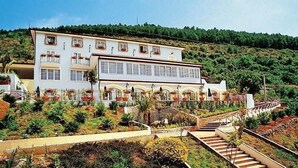 Um edifício grande, com vários andares, fachada simétrica, múltiplas varandas e um jardim ajardinado com diversas plantas e uma escadaria que leva à entrada.