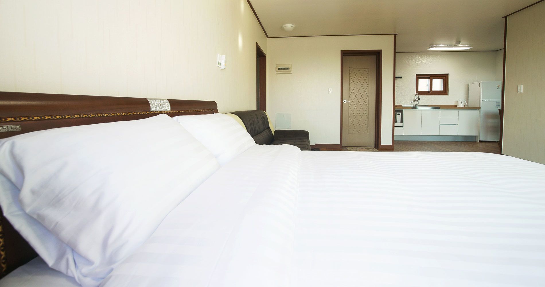 Quarto de hotel com uma cama grande, uma escrivaninha pequena e uma cozinha compacta ao fundo.
