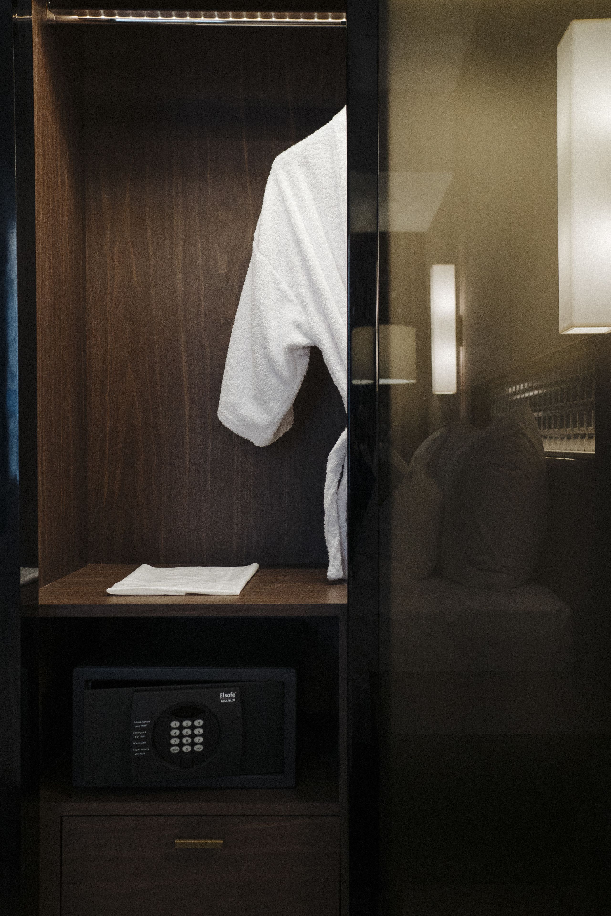 Quarto de hotel com guarda-roupa de madeira, uma toalha branca pendurada na porta, mesa de cabeceira com controle remoto e luminária na parede.