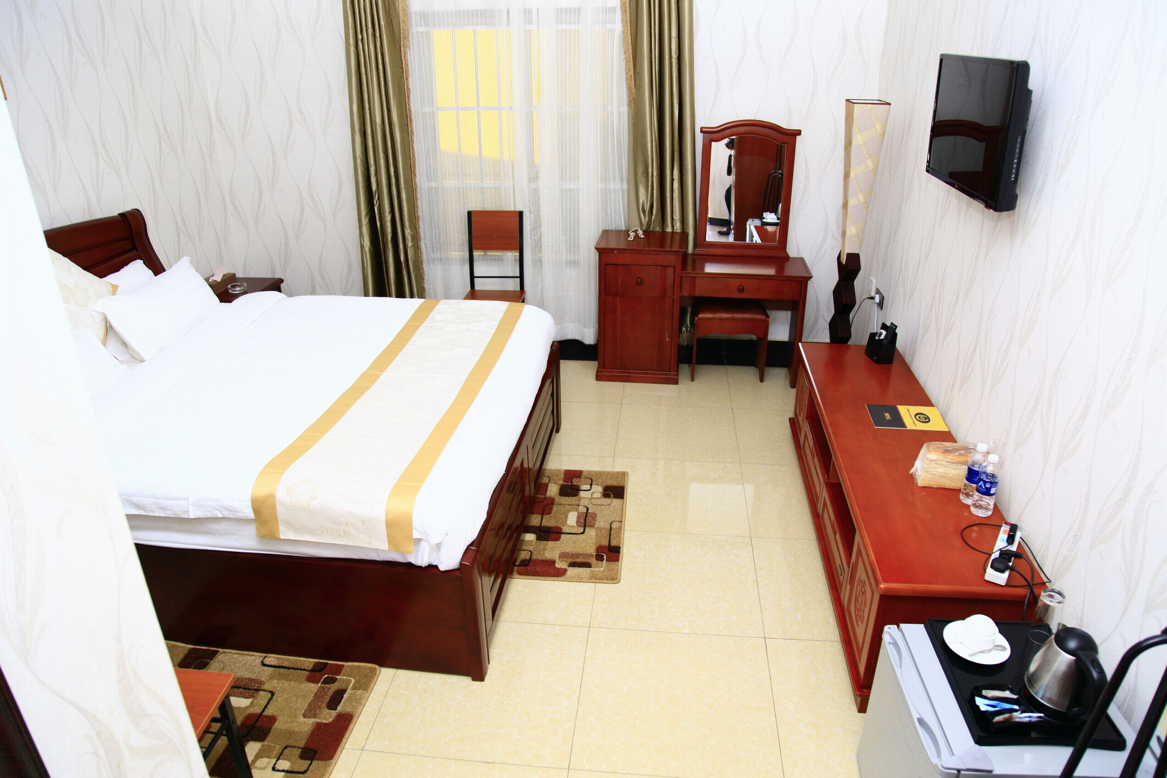 Quarto de hotel com uma cama, uma escrivaninha, uma cadeira, um espelho, uma televisão e uma mesinha com chaleira e chávenas.