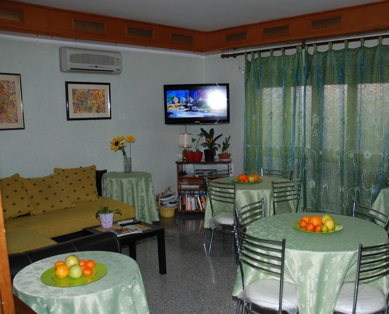 Uma sala de estar com sofá, televisão, mesa de jantar com cadeiras e mesa de centro com frutas.