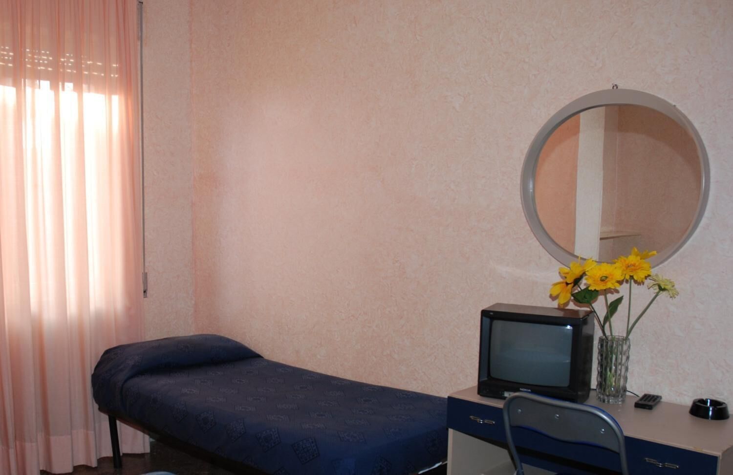 Um quarto pequeno com uma cama de solteiro, uma escrivaninha com computador, uma cadeira, um espelho redondo e um vaso com flores.