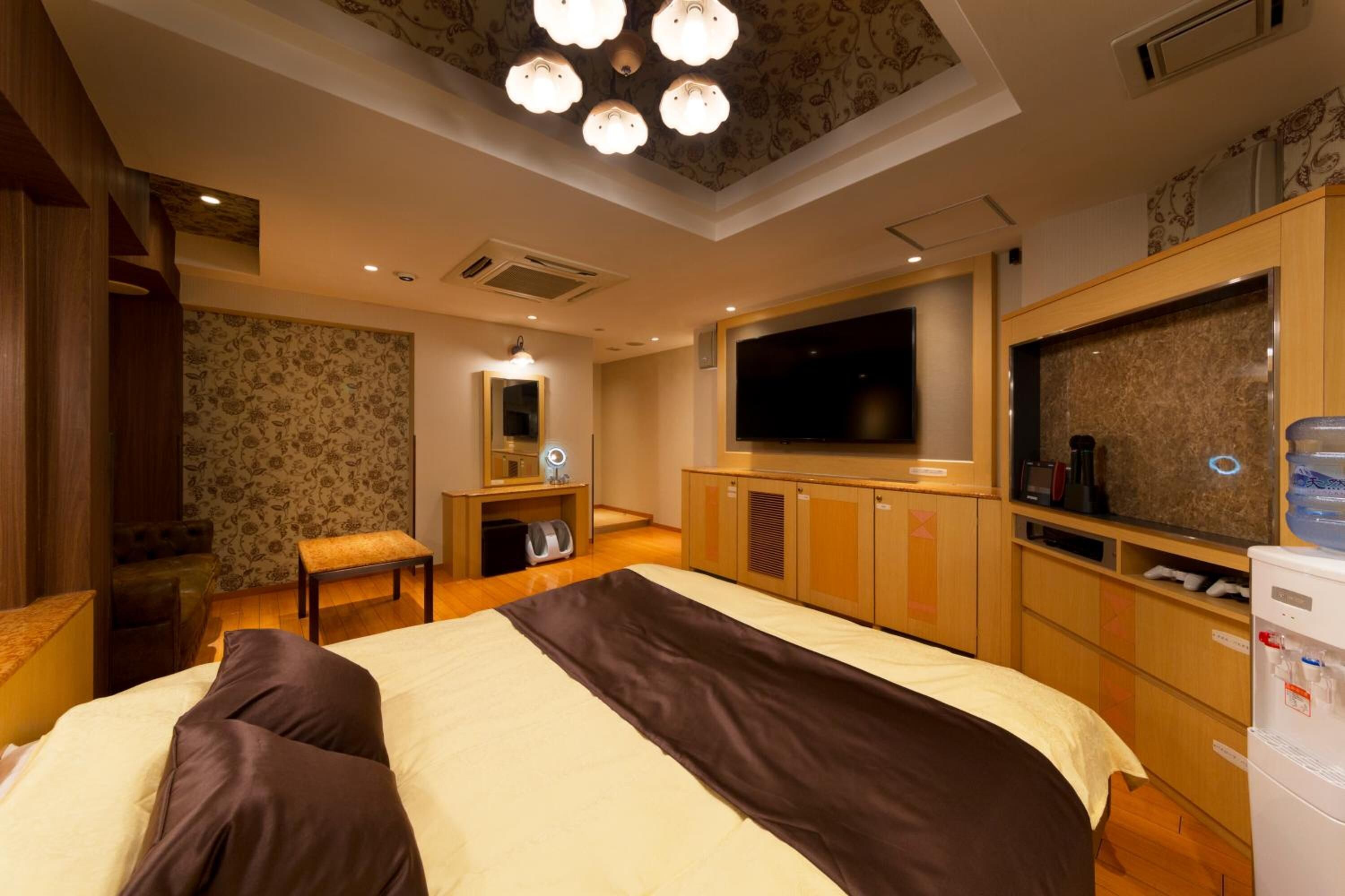 Quarto de hotel com uma cama grande, televisão, dispensador de água e uma pequena mesa com cadeira.