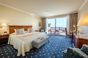 Quarto de hotel com uma cama grande, área de estar com uma poltrona e mesa, e varanda com vista.