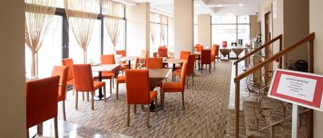 Espaço de refeições de hotel com várias mesas e cadeiras laranja, piso coberto por carpete e grandes janelas com cortinas.