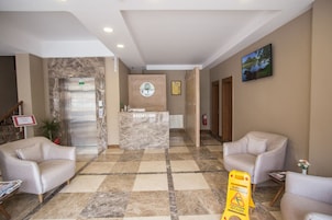 Uma área de recepção com balcão de mármore, duas poltronas, um balcão de atendimento, um elevador e uma placa de aviso no chão.