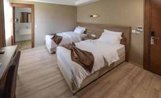 Quarto de hotel com duas camas, piso de madeira, mesa de cabeceira, espelho, escrivaninha pequena e banheiro visível por uma porta aberta.